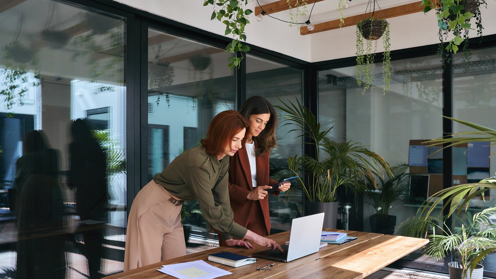 Smiling professional ladies entrepreneurs partners using laptop and tablet technology devices standing at work desk. Two happy business women of young and middle age talking working in green office.